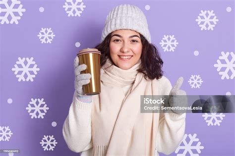 뜨거운 음료와 엄지 손가락으로 열 머그잔을 들고 웃는 젊은 여성 배경에 눈송이와 스튜디오 벽에 포즈 따뜻한 겨울 옷을 입고 여자 좋은 분위기에있는 여성 겨울에 대한 스톡