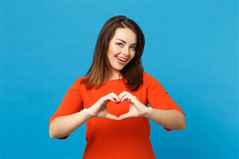 아름 다운 갈색 머리 젊은 여 자가 빨간 오렌지 드레스를 입고 파란 벽 배경 스튜디오 초상화 위에 고립 된 손으로 모양 심장 기호 사람들의 라이프 스타일 패션 컨셉 복사