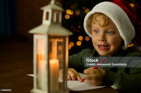 크리스마스 모자를 쓴 소년은 크리스마스 나무에서 등불의 빛에 의해 산타 클로스에게 편지를 씁니다 갈란드 장식품에 대한 스톡 사진