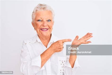 고립 된 흰색 배경 위에 서있는 우아한 셔츠를 입은 선임 회색 머리 의 여성은 손으로 손가락으로 가리키는 동안 카메라에 놀랍고 미소를 지었습니다 가리키기에 대한 스톡 사진