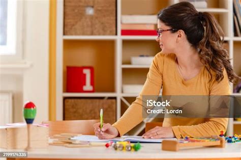 겨자 스웨터를 입은 유치원 교사가 책상에 앉아 따뜻하고 정돈된 교실에서 교육 활동을 준비하면서 공책에 글을 쓰고 있습니다 계획에 대한 스톡 사진 및 기타 이미지 Istock