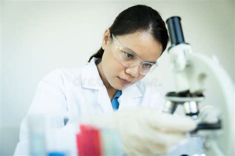 A Woman In A Lab Coat Is Looking Through A Microscope She Is Wearing Safety Goggles And Gloves