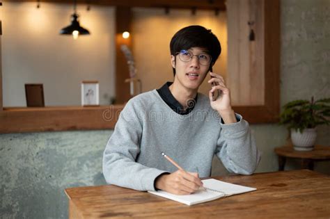 A Young Asian Man Is Talking On The Phone With Someone While Working Remotely At A Coffee Shop