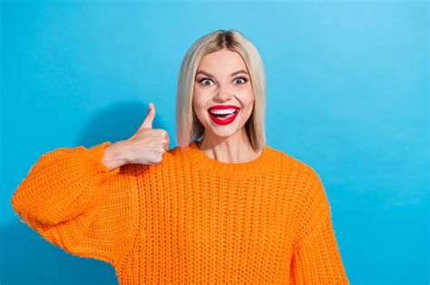 금발 머리를 한 펑키 감동 소녀의 사진은 니트 오렌지 점퍼를 입고 엄지 손가락을 보여주는 파란색 배경에 고립 된 승인을 보여줍니다 Omg에 대한 스톡 사진 및 기타 이미지