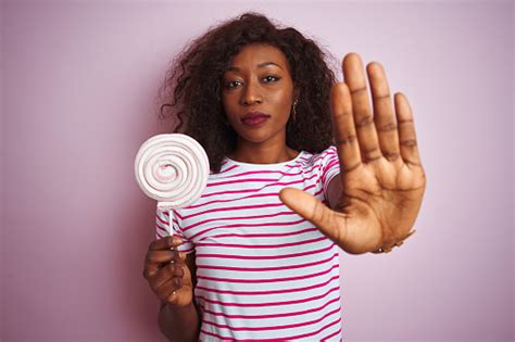 진지하고 자신감있는 표정 방어 제스처로 정지 표지판을하는 열린 손으로 고립 된 분홍색 배경 위에 서있는 달콤한 사탕을 먹는 젊은 아프리카 계 미국인 여성 경계 표지에 대한