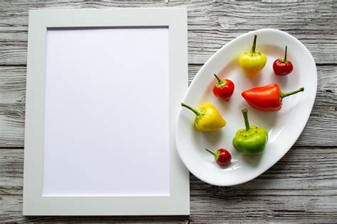 텍스트 또는 그림에 대한 공간이 있는 흰색 프레임입니다 그 옆에는 빨간색 주황색 노란색 및 녹색의 다채로운 작은 칠리 페퍼가있는 흰색 접시가 있습니다 회색 숙성 된 나무의