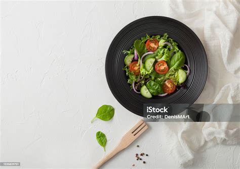 양상추와 토마토 붉은 양파 시금치를 곁들인 신선한 건강한 채식 채소 샐러드와 주걱 포크와 흰색 주방 천이 있는 흰색 배경에 검은 그릇에 새양파와 시금치를 입습니다 빛에 대한