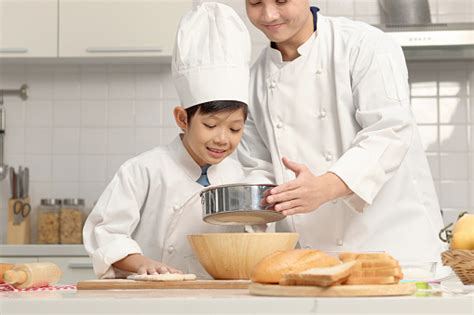 주방에서 모자를 쓰고 요리사 유니폼을 입은 행복한 아시아인 아들과 아버지 귀여운 소년 아이는 아빠가 밀가루를 그릇에 체로 치고 반죽하기 전에 빵 반죽을 준비하고 가족이