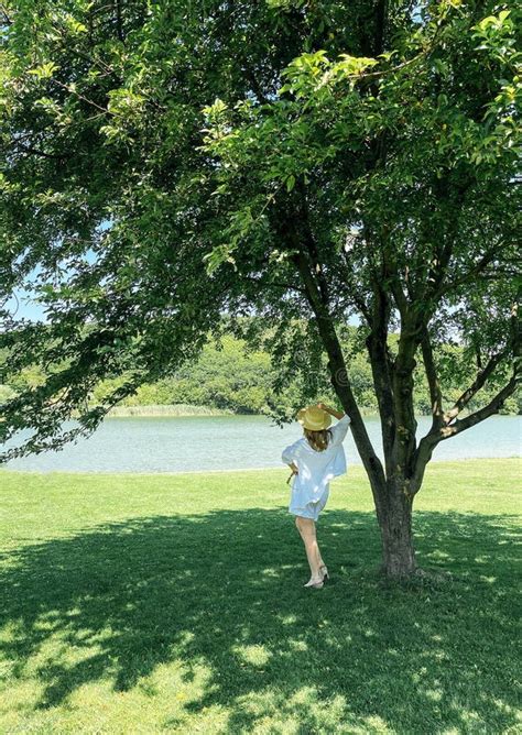 Young Woman In White Clothes And Straw Hat Stands Near Sprawling Tree