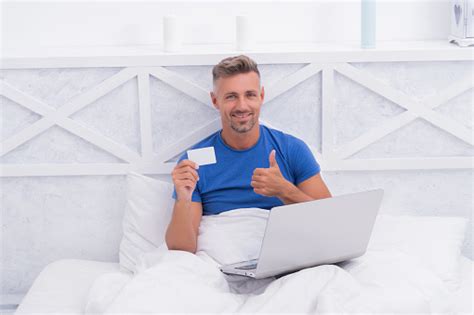 행복한 남자는 침대에서 노트북을 통해 온라인 쇼핑을위한 신용 카드를 들고 엄지 손가락을 제공합니다 전자 뱅킹 고객에 대한 스톡