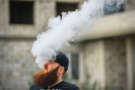 Vape 실제 생활에서 남자 수염 모자와 선글라스 Vaping 흰색 벽돌의 미완성된 건물 맞은편 전자 담배에서에서 큰 수염과 젊은 남자의 초상화 거친에 대한 스톡 사진 및