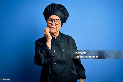 치통이나 치아 질환으로 인해 고통스러운 표정으로 입을 만지는 파란색 배경 위에 밥솥 유니폼과 모자를 쓰고있는 선임 아름다운 회색 머리 요리사 여성 치과 의사 건강관리와