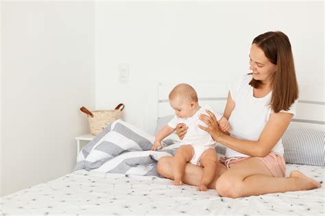 아기와 함께 침실에서 포즈를 취하는 흰색 캐주얼 티셔츠를 입은 매력적인 웃는 여성의 실내 샷 어머니는 딸을 안고 긍정적 인 감정을 표현합니다 2 5 개월에 대한 스톡 사진