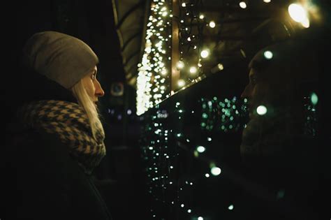 모자를 쓰고 아래 재킷과 볼륨감 있는 스카프를 입은 젊은 금발 여성이 장식된 상점 창문의 화환 카페 클로즈업 창에서 어둠 속에서 보입니다 12월에 대한 스톡 사진 및 기타