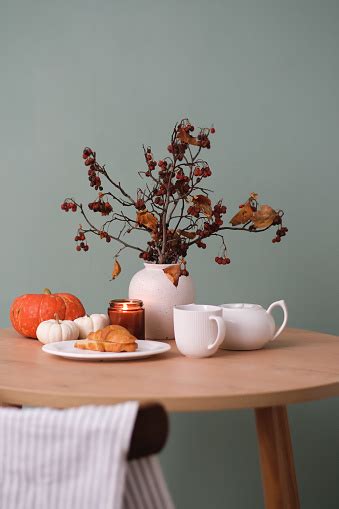 가을 가정 아침 식사 크루아상 마른 열매 가지와 노란 잎이 있는 세라믹 꽃병 나무 테이블과 녹색 벽에 촛불 호박 차 한 잔 찻주전자를 곁들인 접시 0명에 대한 스톡 사진 및