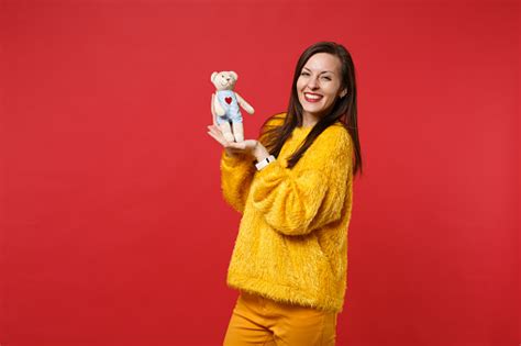 스튜디오에서 밝은 빨간 벽 배경에 고립 된 테 디 베어 봉 제 장난감을 들고 노란색 모피 스웨터에서 즐거운 젊은 여자의 초상화