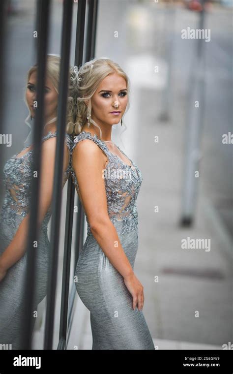 Attractive Blonde Lady Wearing A Long Silver Dress Is Posing Leaning Against The Glass Wall