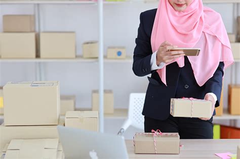 파란 양복을 입은 종교적인 아시아 무슬림 여성이 서서 휴대폰으로 배달하는 패키지 상자의 사진을 찍는다 스타트업 중소기업 프리랜서