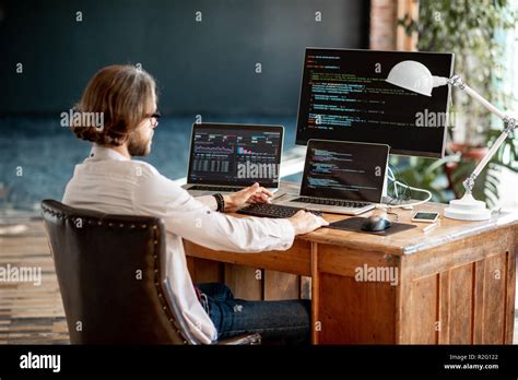 Young Male Programmer Writing A Program Code Sitting At The Workplace With Three Monitors In The