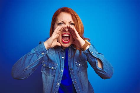 파란색 고립 된 배경 위에 서있는 데님 셔츠를 입은 젊은 아름다운 빨간 머리 여성은 입에 손을 얹고 큰 소리로 화를 냅니다 데님에 대한 스톡 사진 및 기타 이미지 Istock