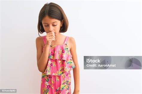 고립 된 흰색 배경 위에 서있는 분홍색 꽃 무늬 드레스를 입은 어린 아름다운 소녀는 감기 또는 기관지염의 증상으로 기분이 좋지 않고 기침합니다 헬스케어 개념 기침하기에 대한