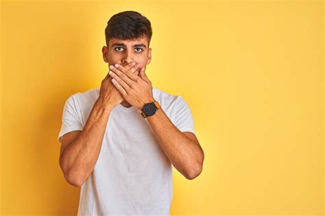 고립 된 노란색 배경 위에 서있는 흰색 티셔츠를 입은 젊은 인도 남자는 실수로 손으로 입을 덮는 충격을 받았습니다 비밀 개념입니다 T 셔츠에 대한 스톡 사진 및 기타 이미지