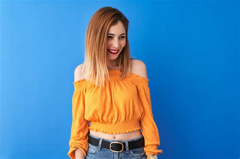 주황색 캐주얼 티셔츠를 입은 아름다운 빨간 머리 여자는 얼굴에 미소와 함께 옆으로 찾고 고립 된 파란색 배경 위에 서 자연스러운 표정 자신감 있게 웃고 있습니다 T 셔츠에