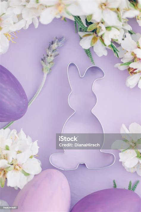 흰색 빈티지 액자에 파스텔 색상의 보라색 표면에 아몬드 꽃으로 둘러싸인 분홍색과 보라색 부활절 달걀과 라벤더가 있는 쿠키 커터 부활절 토끼 모양의 아름답고 섬세한 클로즈업