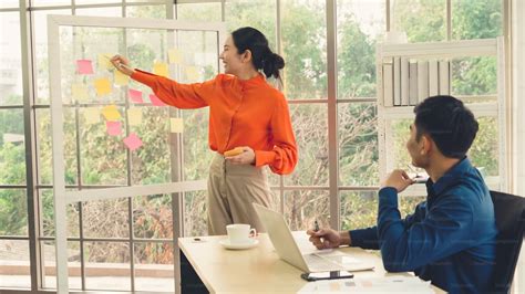 비즈니스 사람들은 사무실의 프로젝트 계획 보드에서 일하고 동료 친구와 대화하여 프로젝트 개발을 분석합니다 그들은 유리 벽에 게시 된 스티커 메모를 사용하여 정리합니다