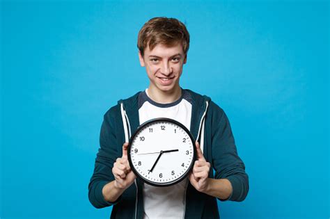 스튜디오에서 파란색 벽 배경에 고립 된 라운드 시계를 들고 캐주얼 의류에 웃는 젊은 남자의 초상화 시간이 부족 합니다 사람들은 진심으로 감정 라이프 스타일 개념입니다 복사