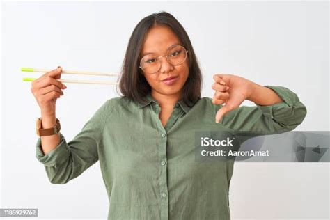 화가 난 얼굴로 고립된 흰색 배경 위에 젓가락을 들고 안경을 쓰고 있는 젊은 중국 여성 엄지손가락으로 싫어하는 것을 보여주는 부정적인 표지판 거부 개념 개념에 대한 스톡