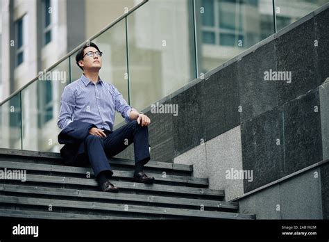 Tired Serious Asian Businessman Sitting On Steps And Looking Up In The