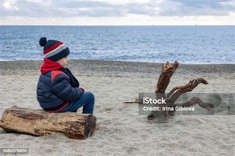 겨울 재킷을 입고 니트 모자를 쓴 귀여운 소년이 걸림돌 옆 통나무에 황량한 해변에 뒤에서 앉아 있습니다 6 7 살에 대한 스톡 사진 및 기타 이미지 Istock