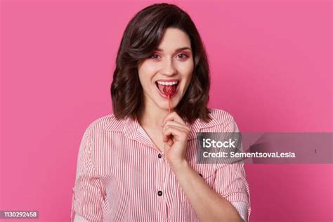 아름 다운 여성 모델의 사진 입에 사탕을 보유 하 고 여가 시간을 즐기고 줄무늬 셔츠를 입고 카메라에서 직접 분홍색 배경 감싸고 달콤한 사탕에 포즈 핀 업 소녀는 미소 및