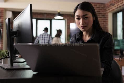 asian software developer coming at desk and sitting down holding laptop with coding interface