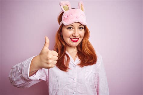 젊은 아름 다운 빨간 머리 여자 핑크 고립 된 배경 위에 잠 옷을 입고 행복 한 엄지 손가락 을 하 고 손제스처 성공을 보여주는 카메라를 보고 표정을 승인합니다 긍정적인