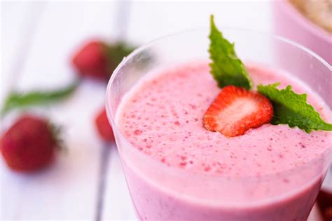 민트와 딸기로 장식된 유리에 딸기와 우유를 곁들인 칵테일 근처에는 헤라클레스 플레이크가 있는 분홍색 컵과 하얀 나무 배경에 느슨한 딸기가 있습니다 아침 여름 아침 식사