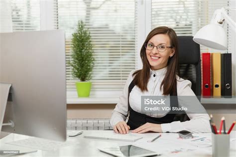 카메라 보면서 아름 다운 웃는 브라운 헤어 비즈니스 여자 양복과 태블릿 책상에 앉아 안경 빛 사무실에서 문서와 현대 모니터