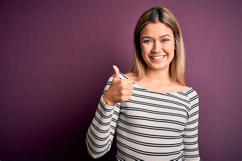 보라색 고립 된 배경 위에 캐주얼 스트라이프 셔츠를 입고 젊은 아름다운 금발의 여자는 손으로 제스처를 행복 엄지 손가락을하고 성공을 보여주는 카메라를 보고 표정을 승인합니다