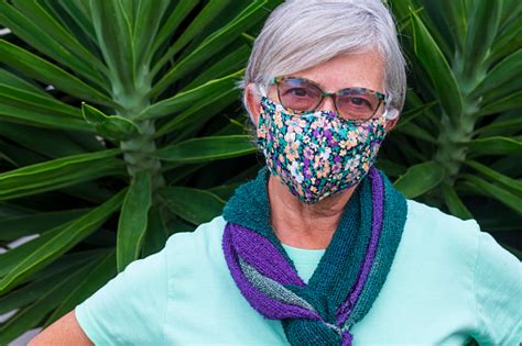 코로나 바이러스 코로나바이러스 전염병으로 인해 꽃이 만발한 수술용 마스크를 쓰고 카메라를 바라보는 고위 미소 여성의 초상화 녹색 열대 식물의 배경 70 79세에 대한 스톡