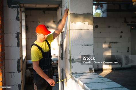 건설 현장의 건설 작업자는 테이프 측정으로 창 개구부 및 벽돌 벽의 길이를 측정합니다 코티지는 다공성 콘크리트 블록 작업복으로 만들어졌습니다 점프수트와 야구 모자 30 34