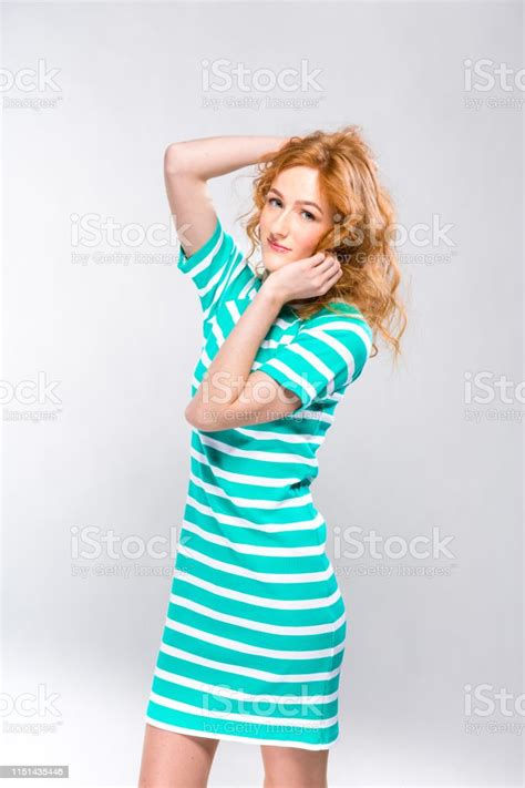 회색 배경에 스튜디오에서 파란색 줄무늬가 있는 여름 드레스에 빨간 곱슬 머리를 가진 젊고 아름 다운 여자의 무릎 초상화 여름 방학 관광 및 여름 옷의 테마 관광객에 대한