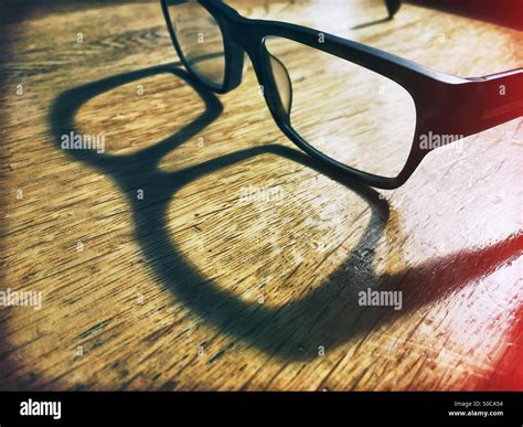 A Pair Of Thick Rimmed Reading Glasses Sitting On A Wooden Desk Casting A Shadow In Sunlight