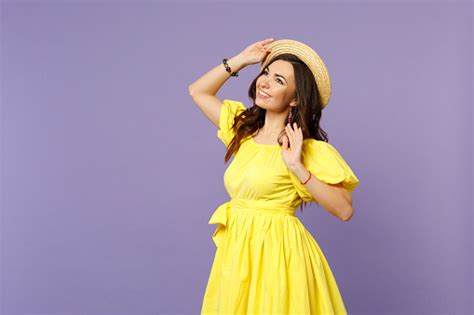 파스텔 바이올렛 벽 배경에 고립 된 찾고 여름 모자에 손을 유지 노란 드레스에 잠겨있는 웃는 젊은 여자의 초상화 사람들은 진심으로 감정 라이프 스타일 개념입니다 복사 공간을