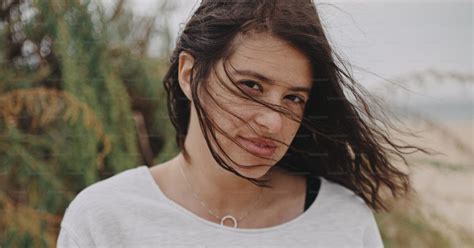 바람이 부는 머리카락과 스웨터를 입고 푸른 잔디와 바다를 배경으로 모래 해변에 앉아 고요한 순간을 가진 아름다운 평온한 여성 해안에서 휴식을 취하는 세련된 젊은 여성