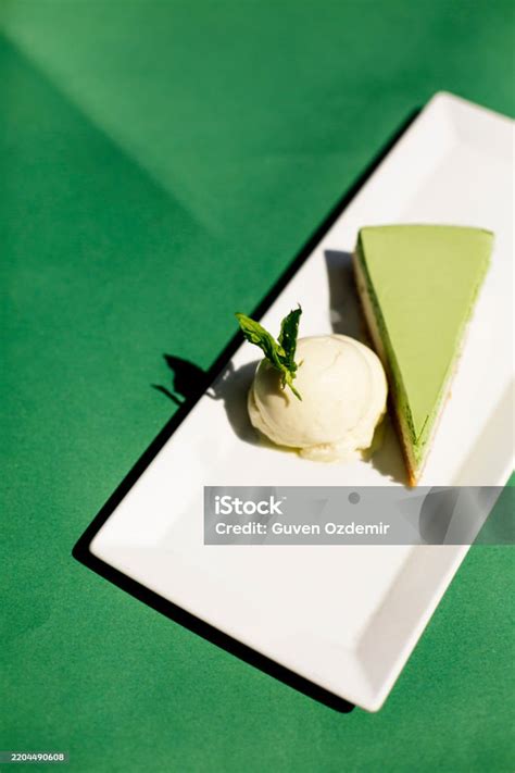 흰색 직사각형 접시에 바닐라 아이스크림 한 스쿱을 곁들인 녹차 말차 치즈케이크 슬라이스의 평면도 얼룩덜룩한 햇빛 디저트 및 제과점 컨셉의 녹색 테이블 위에 놓여 있습니다 0