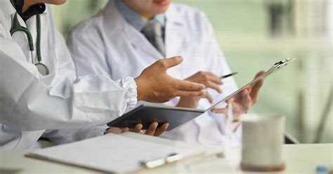 컴퓨터 태블릿과 클립보드를 들고 의사로 일하는 두 명의 똑똑한 남자가 질서 정연한 직장을 배경으로 의사 작업 책상에 함께 앉아 토론하고 앉아 있는 잘린 이미지 사진