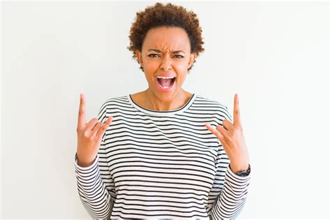 젊은 아름 다운 아프리카 계 미국인 여자 흰색 배경 에 줄무늬 스웨터를 입고 미친 표정으로 외치고 바위 기호를 하 고 손을 위로 음악 스타 무거운 개념 개념에 대한 스톡