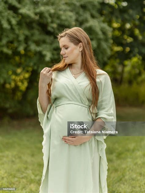 백인 임산부는 머리카락을 잡고 바라보고 배에 손을 얹고 공원에 서 있다 아기를 기대하는 동안 의료 및 미용 개념 긴 머리 여성 갈색 머리는 녹색 드레스를 입는다 수직 임신에