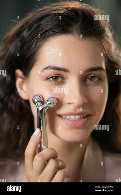 Gorgeous Young Brunette Woman Using Metallic Massage Roller Upon Her Face And Looking At You
